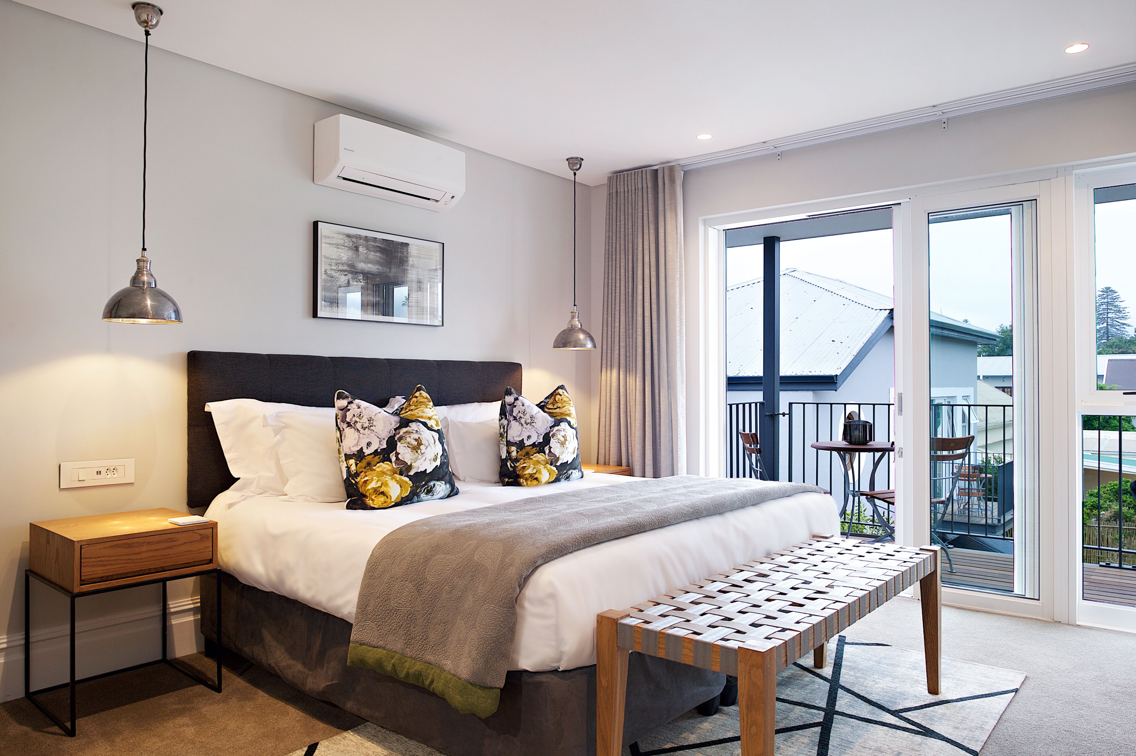 Quarto moderno com uma cama grande, mesas de cabeceira, um banquinho e uma varanda com vista.