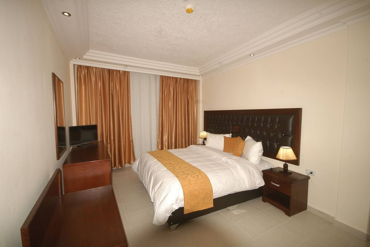 Quarto de hotel com uma cama grande, uma TV em um suporte de madeira, uma mesa de cabeceira com uma luminária e um espelho na parede.