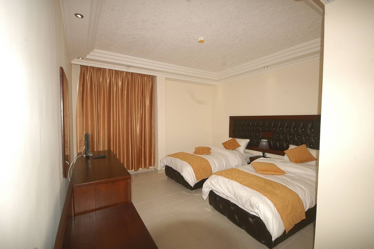 Quarto de hotel com duas camas, uma escrivaninha de madeira e uma janela grande com cortinas.
