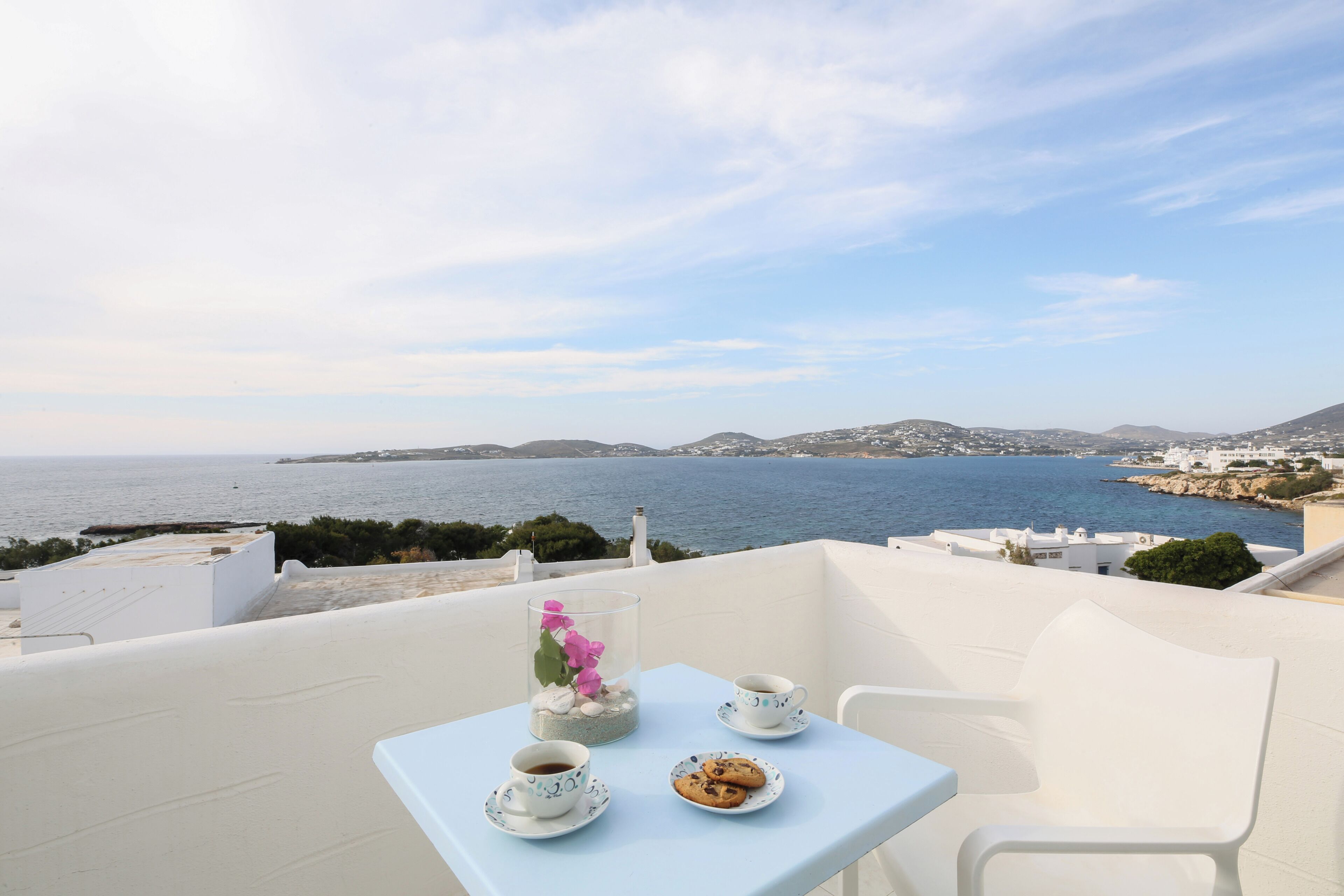 Uma mesa com uma xícara de café, um prato de biscoitos e um vaso de vidro com flores em uma varanda com vista para o mar.