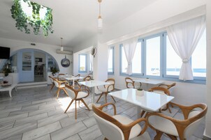 Quarto claro com paredes brancas, grandes janelas e vista para o mar. Há mesas e cadeiras dispostas para refeições.