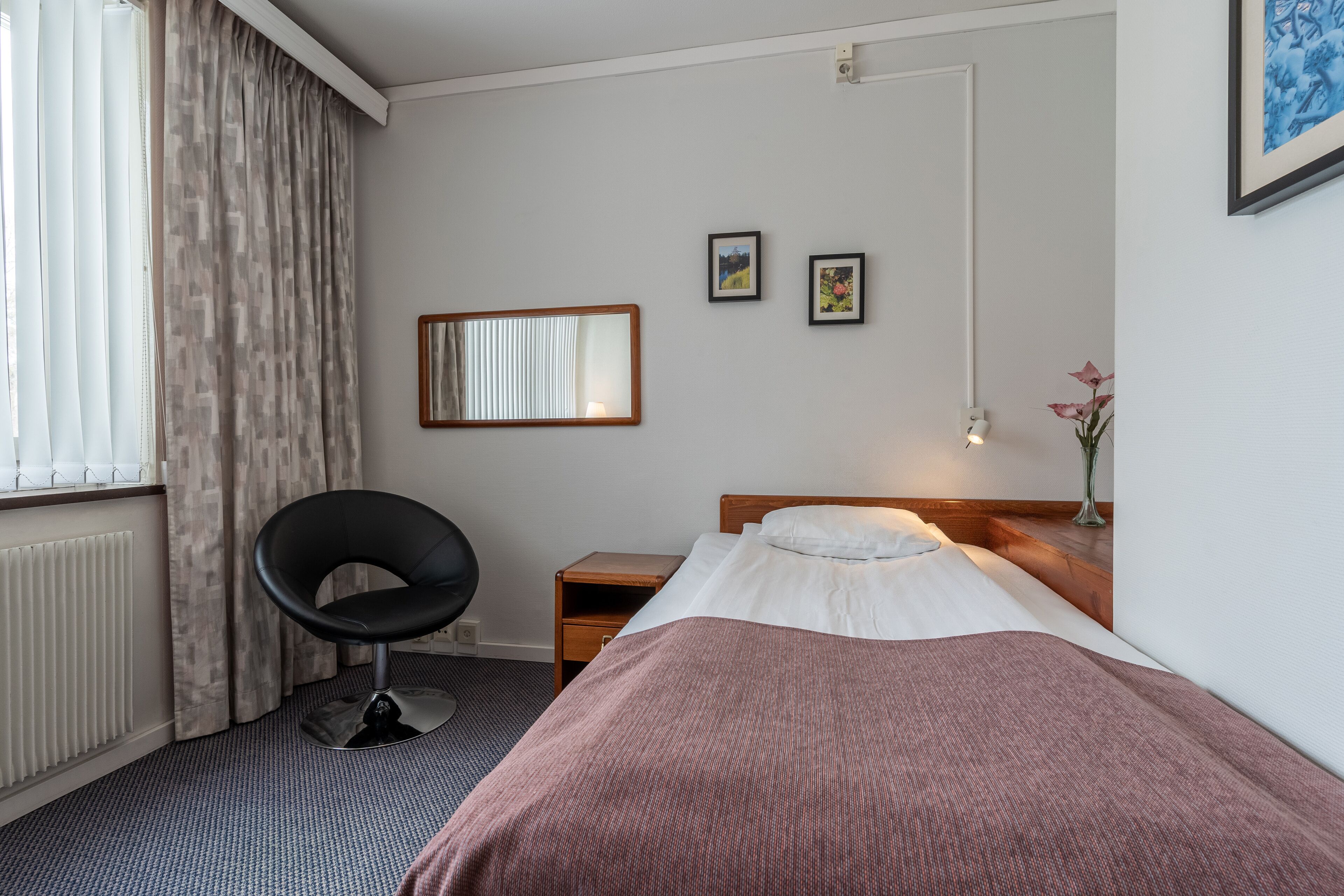 Quarto de hotel com uma cama, uma cadeira, uma mesa de cabeceira, um espelho e uma janela com cortinas.