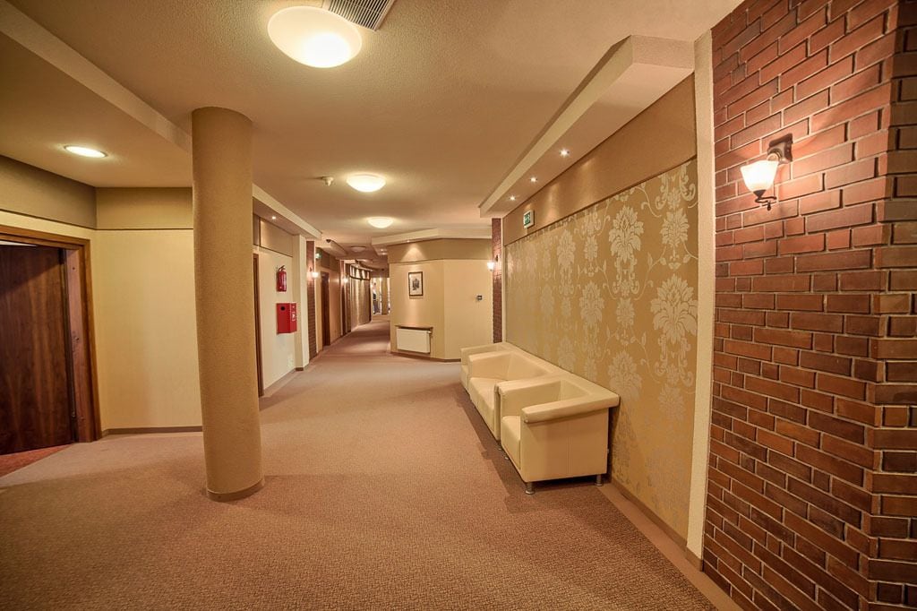 Um corredor de hotel com piso em carpete, parede de tijolos com papel de parede estampado, um sofá bege e um pilar.
