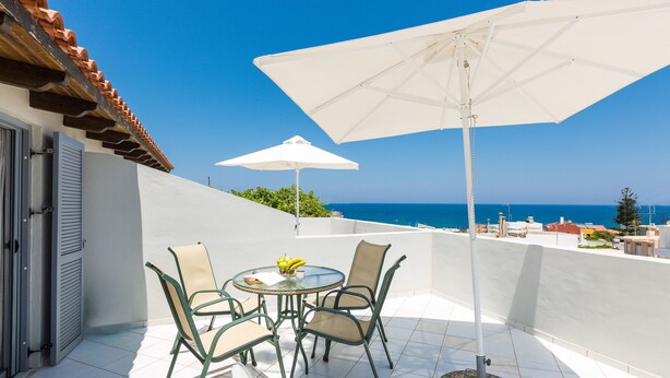 Um terraço com mesa, cadeiras e dois guarda-sóis brancos, com vista para o mar.