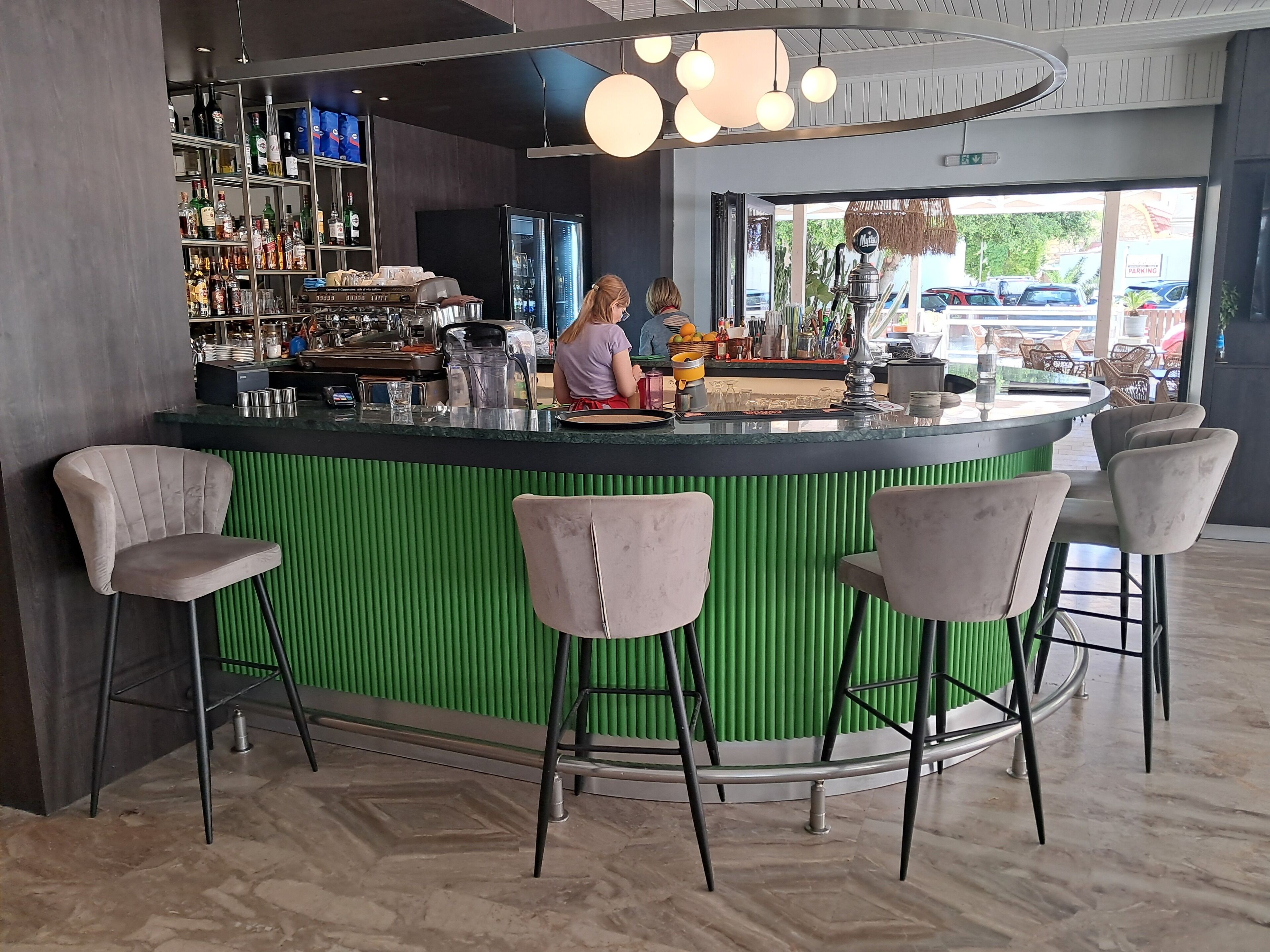 Uma área de bar com balcão verde, banquetas e vista para carros estacionados lá fora.