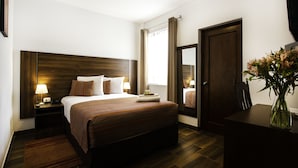 Quarto de hotel com uma cama grande, luminárias de cabeceira, uma cabeceira de madeira, um espelho, uma mesinha de cabeceira de madeira, uma pequena mesa com um vaso de flores e um tapete marrom.