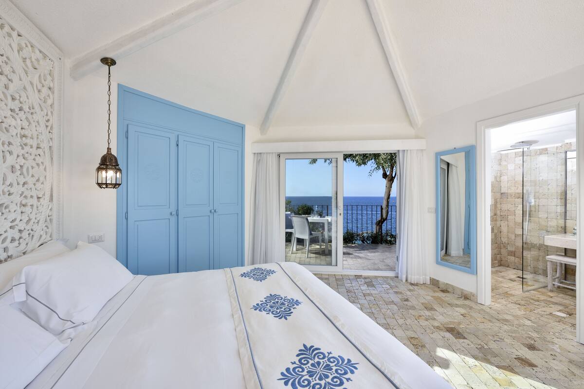 Quarto com cama, porta azul, varanda com vista para o mar e banheiro com chuveiro de vidro.