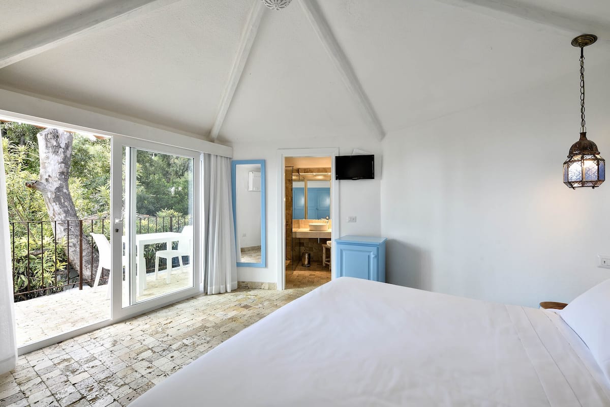 Quarto com cama grande, TV fixada na parede e porta de vidro deslizante para uma varanda com móveis de exterior.