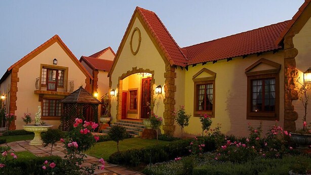 Uma casa tradicional com telhado de telhas vermelhas, pilares de pedra e um jardim bem cuidado com flores cor-de-rosa.