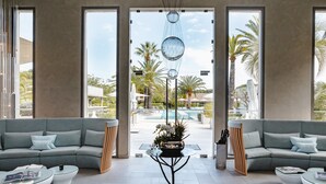 Uma área de estar moderna com dois sofás, uma mesa central e amplas janelas com vista para uma piscina e palmeiras.