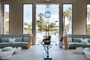 Uma área de estar moderna com dois sofás, uma mesa central e amplas janelas com vista para uma piscina e palmeiras.