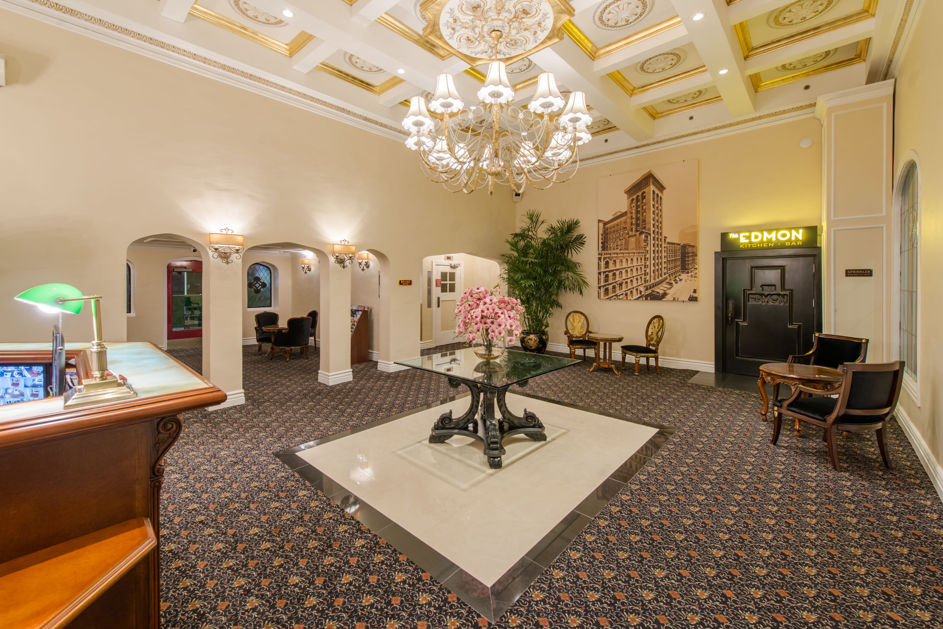 Um hall de hotel com um candelabro, um balcão de recepção, uma mesa com flores e uma placa com 'The Edmon'.