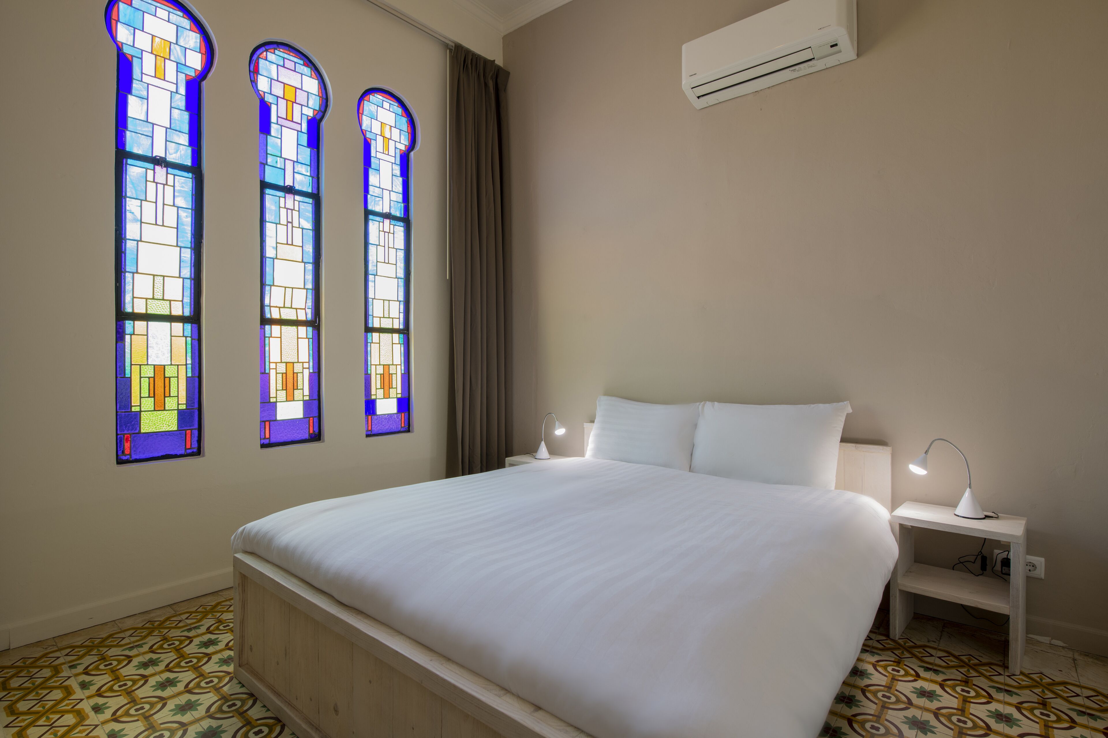Quarto com uma cama grande, mesa de cabeceira, abajur e três janelas de vitrais.