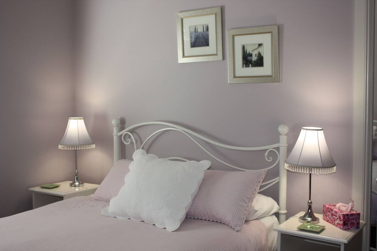Cama bem arrumada com roupa de cama branca e rosa, duas mesas de cabeceira com abajures e duas fotos emolduradas na parede.