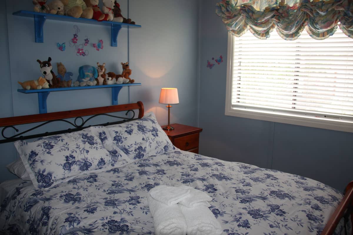 Quarto com colcha florida, cabeceira de madeira, mesa de cabeceira com abajur e prateleiras com bichos de pelúcia.