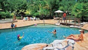 Piscina com pessoas relaxando e nadando, cercada por palmeiras e espreguiçadeiras.