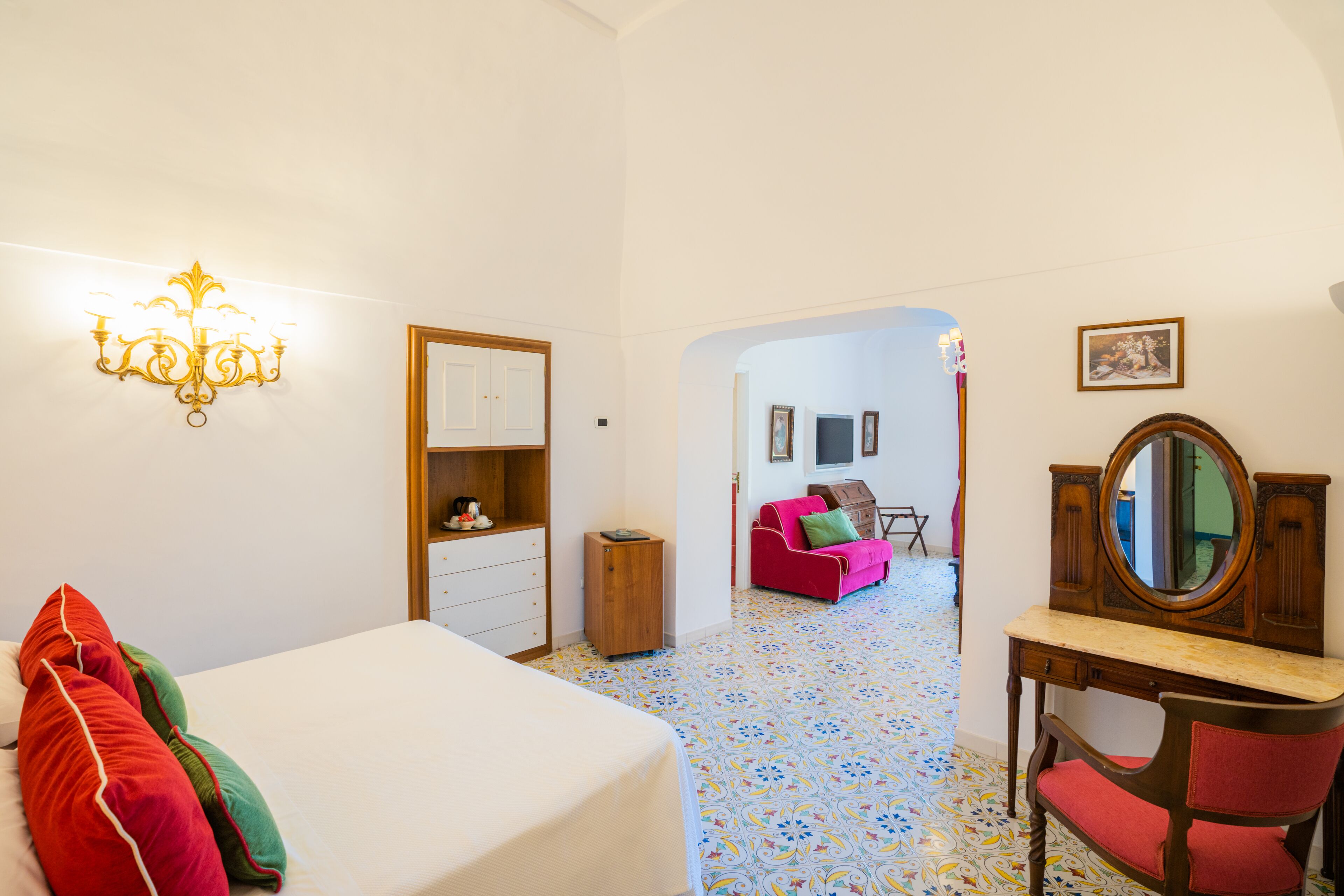 Quarto com cama, cômoda, penteadeira com espelho e piso estampado em cores vibrantes.