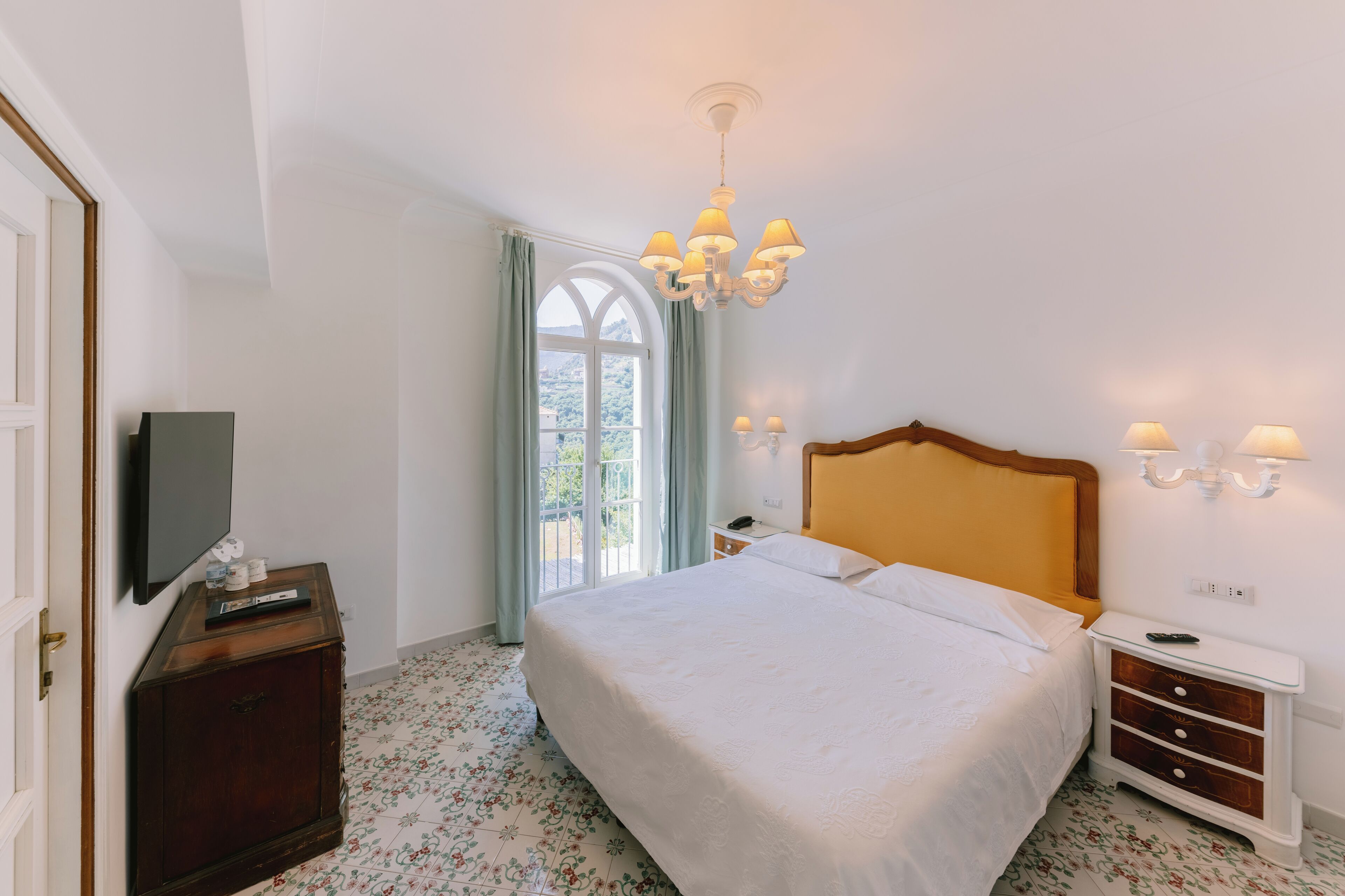 Quarto com cama, televisão sobre um móvel de madeira, um candelabro e janela com cortinas.