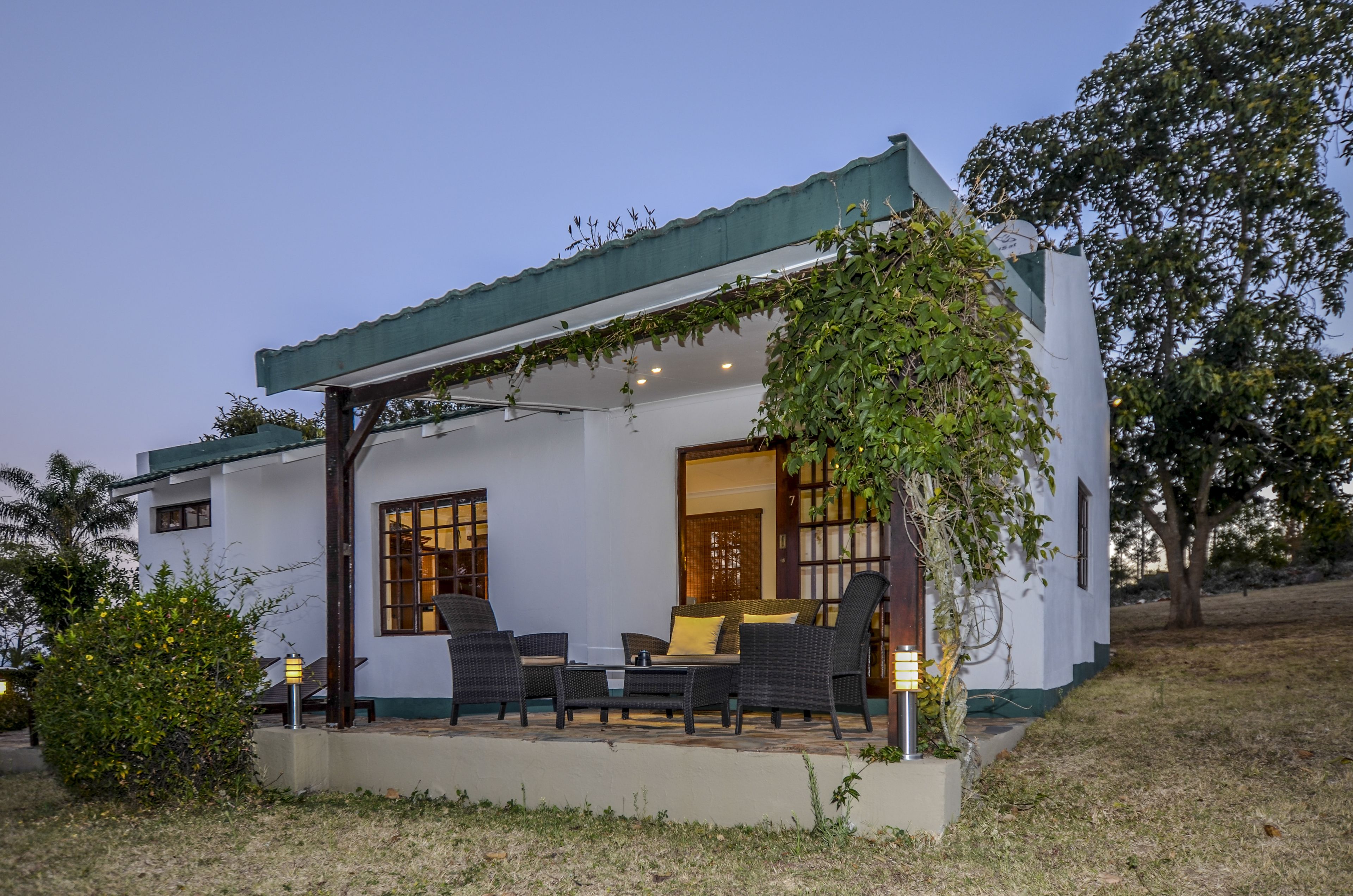 Um edifício branco com telhado verde, um alpendre coberto com mobiliário de vime e uma pequena área de jardim.