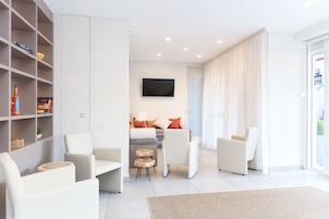 Uma sala de estar moderna com móveis brancos, uma televisão de tela plana e uma janela grande com cortinas.