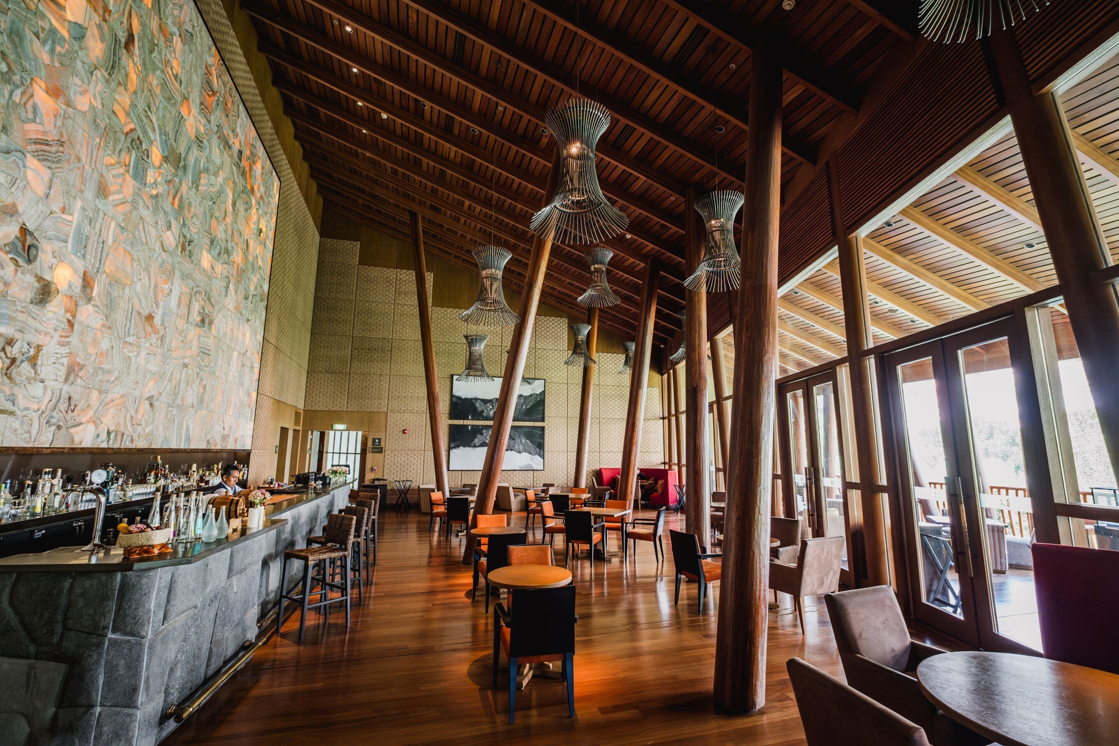 Um espaço amplo de bar com balcão de pedra, teto de madeira e grandes janelas que permitem entrada de luz natural.
