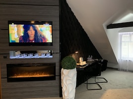 Uma sala de estar moderna com televisão de tela plana na parede, lareira, área de jantar com mesa e cadeiras, e uma janela grande que proporciona luz natural.