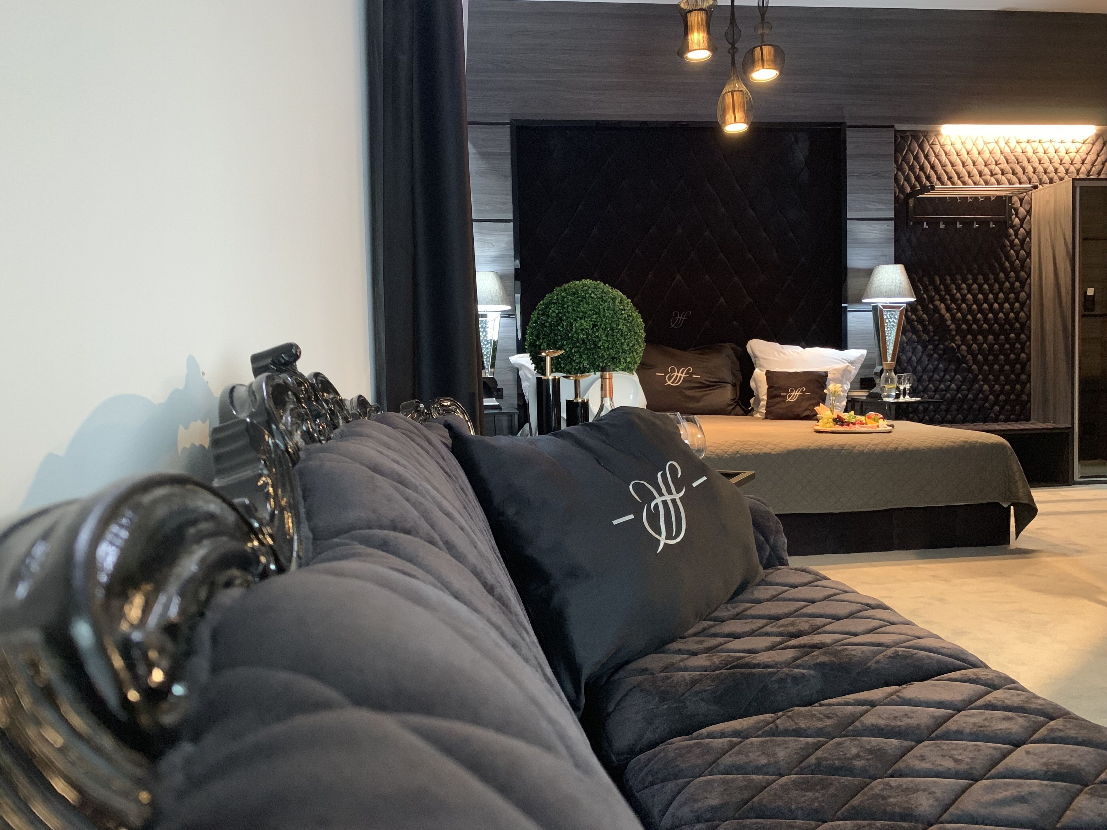 Quarto moderno com esquema de cores escuras, cama grande com cabeceira acolchoada, mesa redonda pequena com abajur e planta decorativa.