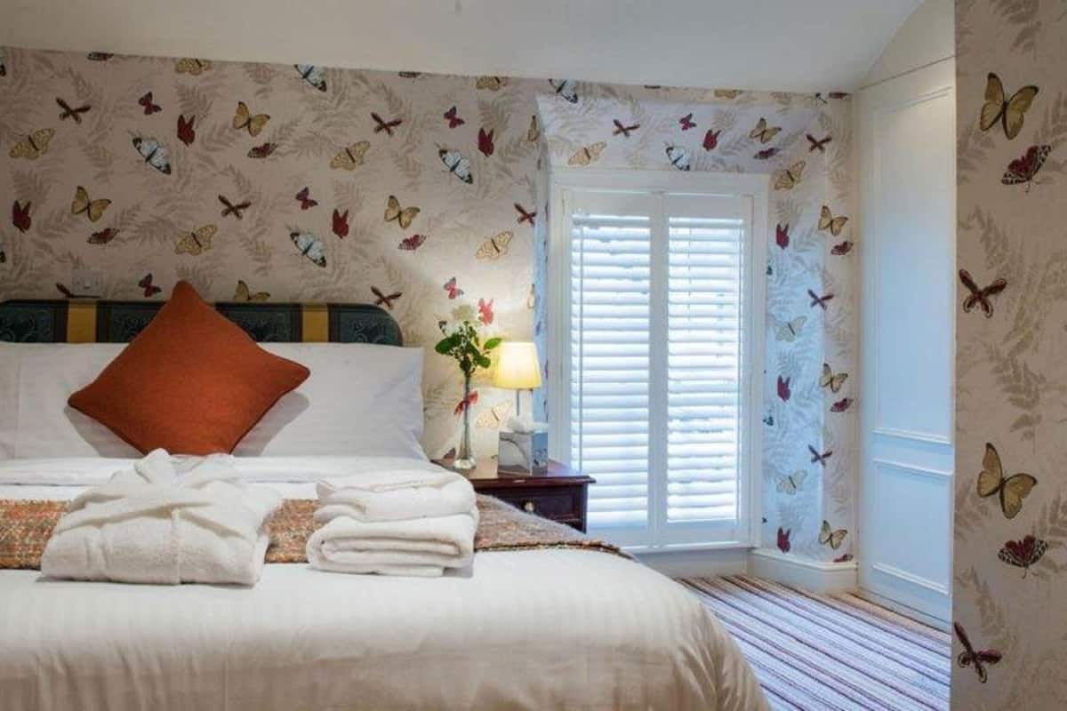 Uma cama bem arrumada com colcha de padrão floral, um travesseiro marrom e duas toalhas brancas dobradas. Uma mesa de cabeceira com luminária e um vaso de flores está ao lado da cama. O quarto tem papel de parede com padrão de borboletas e uma janela com persianas.