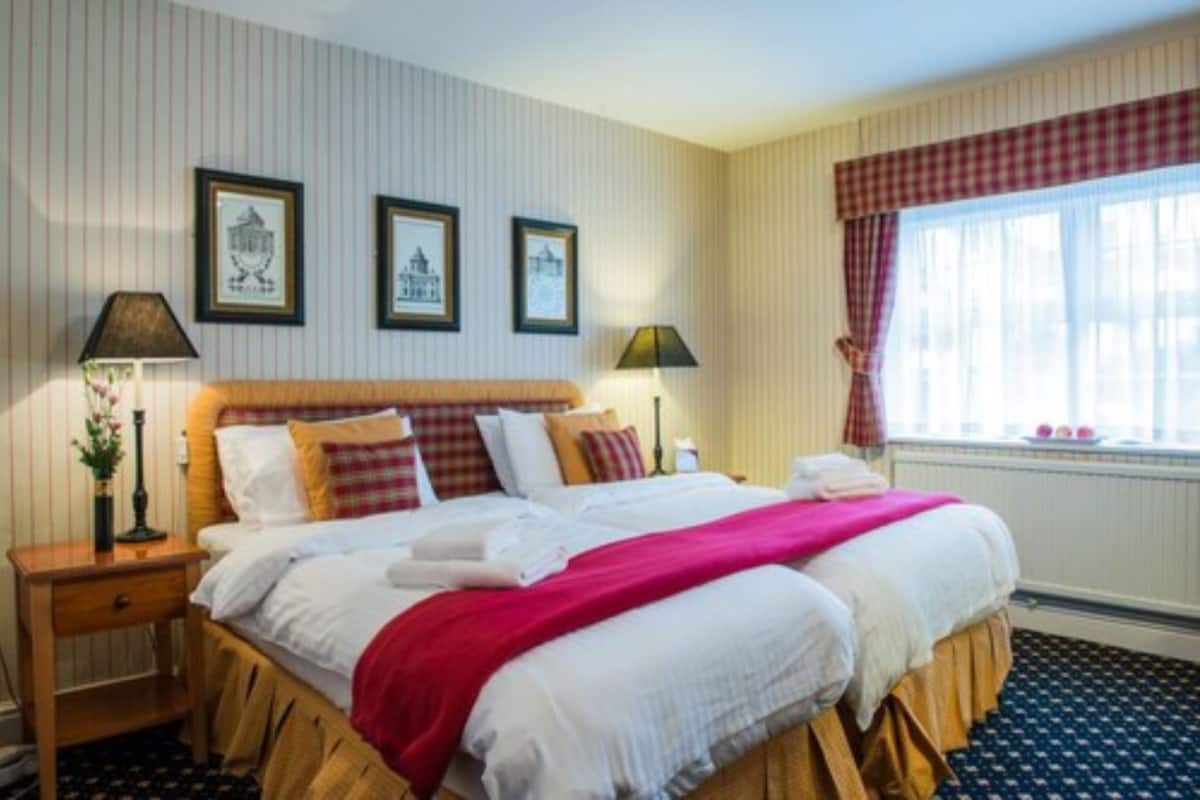 Quarto de hotel com uma cama grande, duas mesas de cabeceira, uma janela com cortinas e quadros nas paredes.