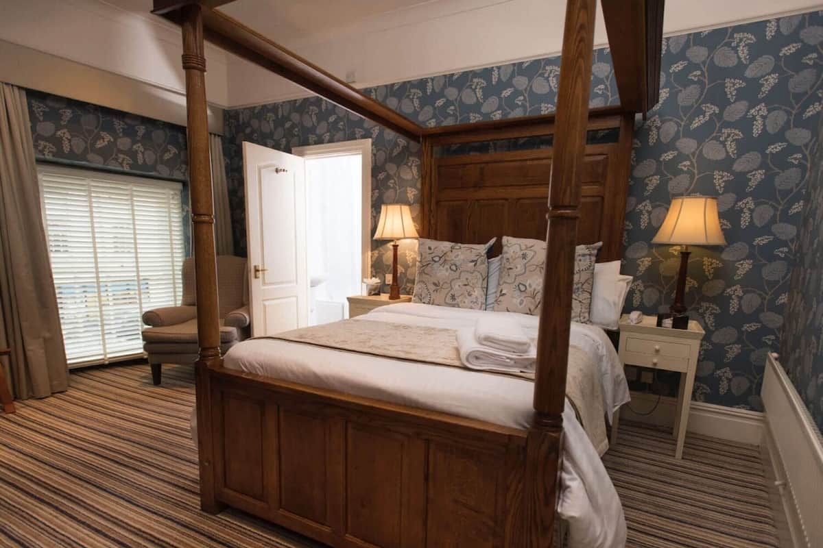 Cama com dossel e estrutura de madeira, com cabeceira branca e colcha estampada com flores. Há uma mesa de cabeceira com abajur e telefone, e uma cadeira no canto. O quarto tem papel de parede azul com padrão de folhas e uma janela com persianas.