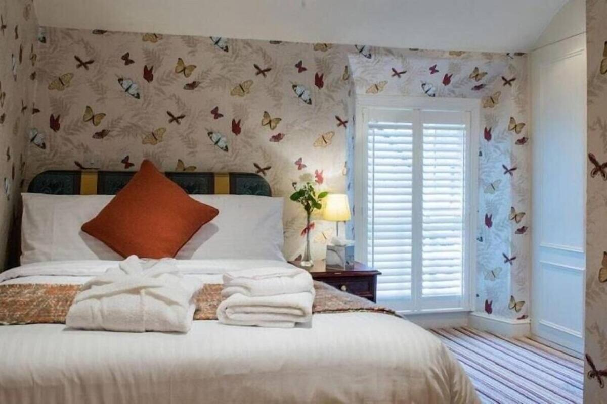 Quarto com cama, almofadas, mesa de cabeceira, abajur e papel de parede com padrão de borboletas.