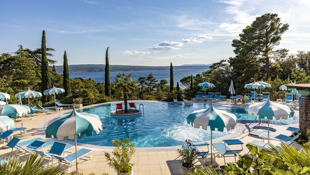 Área de piscina com espreguiçadeiras e guarda-sóis, cercada por árvores e vista para o mar.