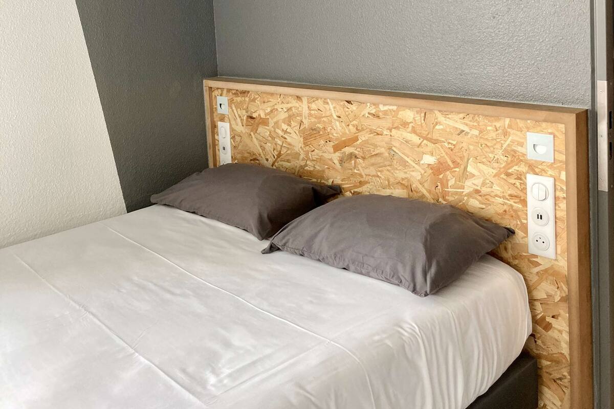 Uma cama com cabeceira de madeira, duas almofadas e um colchão branco num quarto com paredes cinzentas e teto inclinado.
