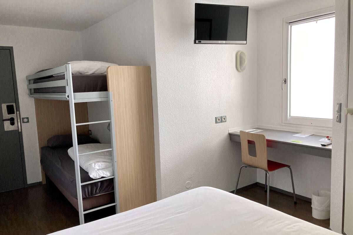 Uma cama de solteiro, uma escrivaninha com cadeira, uma televisão e uma janela em um quarto de hotel.
