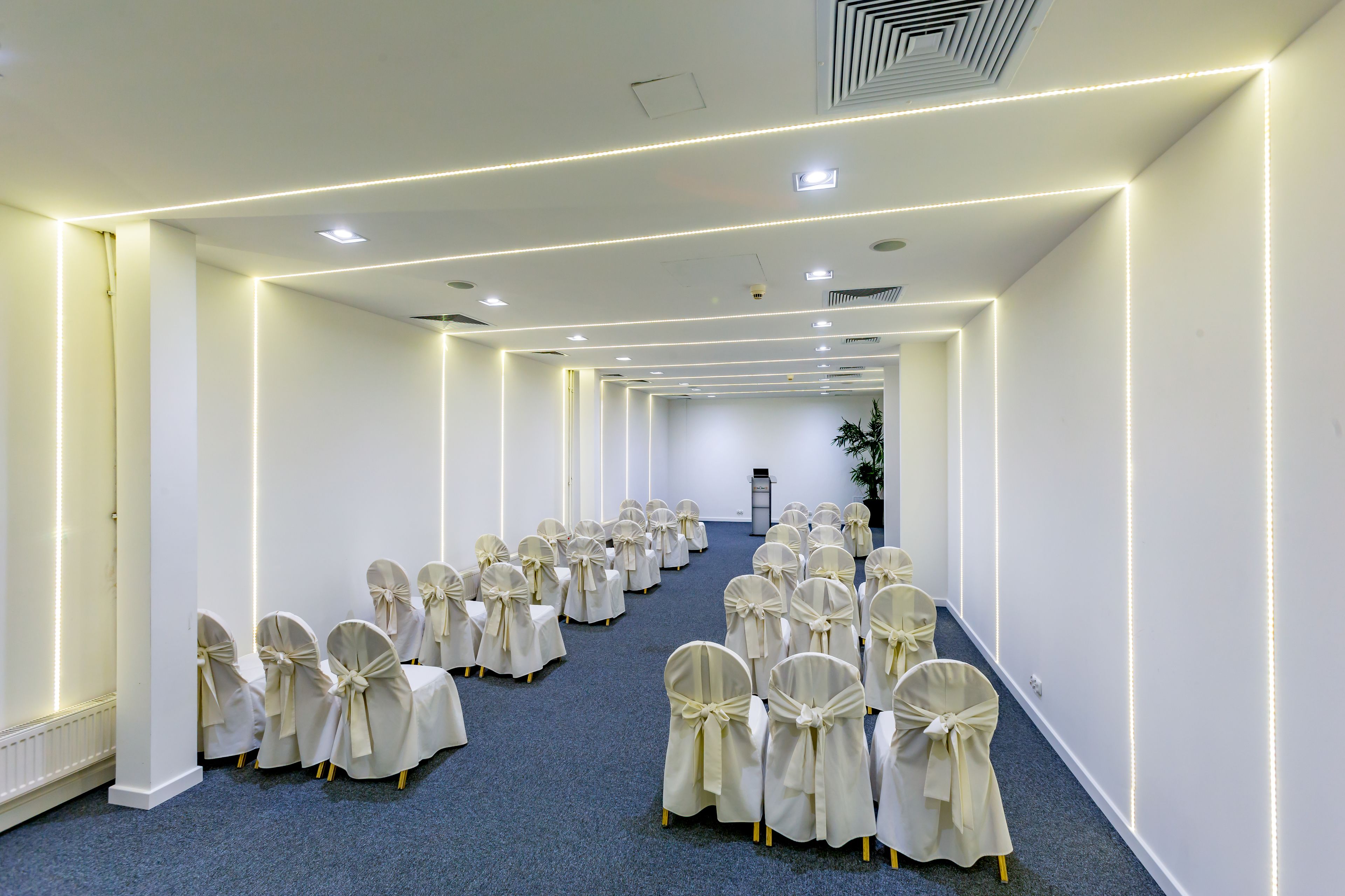 Um corredor de hotel com fileiras de cadeiras e mesas, paredes brancas e luminárias no teto.