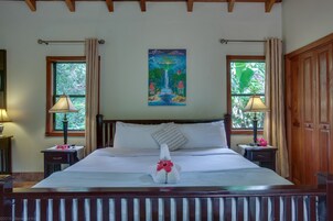 Uma cama bem arrumada com uma peça central decorativa em formato de cisne, uma pintura de uma paisagem tropical acima dela e duas luminárias de cada lado da cama.