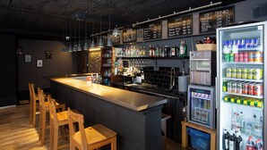 Uma área de bar com um balcão comprido, vários bancos, uma geladeira repleta de bebidas e um quadro de preços de bebidas.
