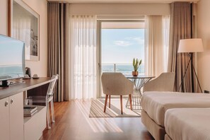 Quarto de hotel com cama, escrivaninha, cadeira, televisão, luminária e vista para o oceano.