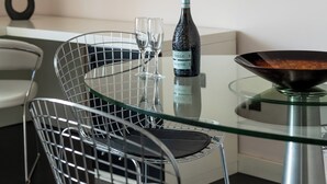 Uma sala de jantar moderna com mesa de vidro, cadeiras de metal, uma garrafa de vinho e uma tigela sobre um suporte de vidro.