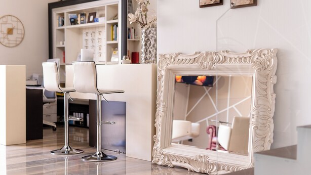 Interior moderno com um espelho branco ricamente decorado, dois banquetes e uma estante de livros ao fundo.