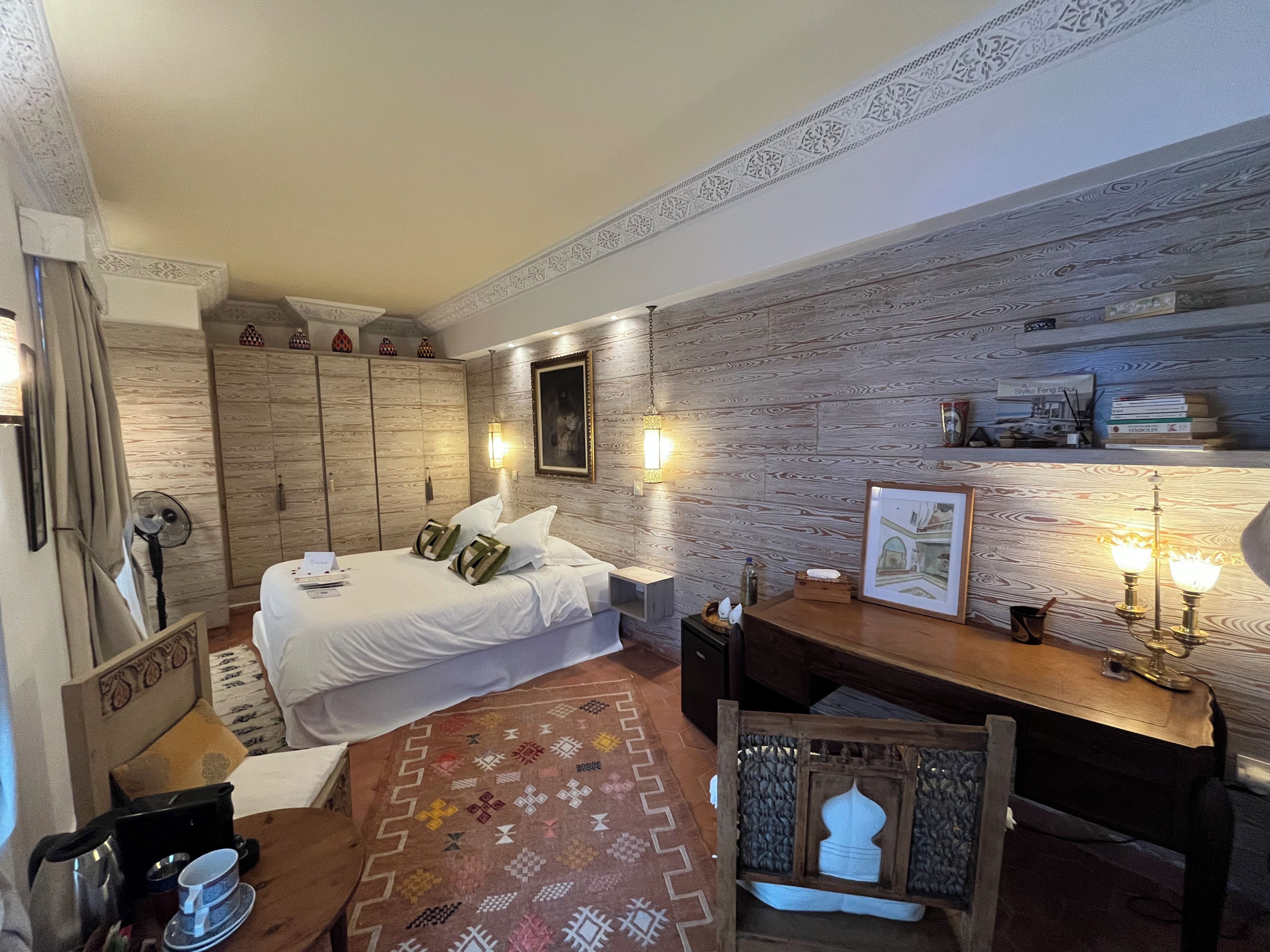 Quarto com cama, mesa de cabeceira, abajur e tapete estampado.