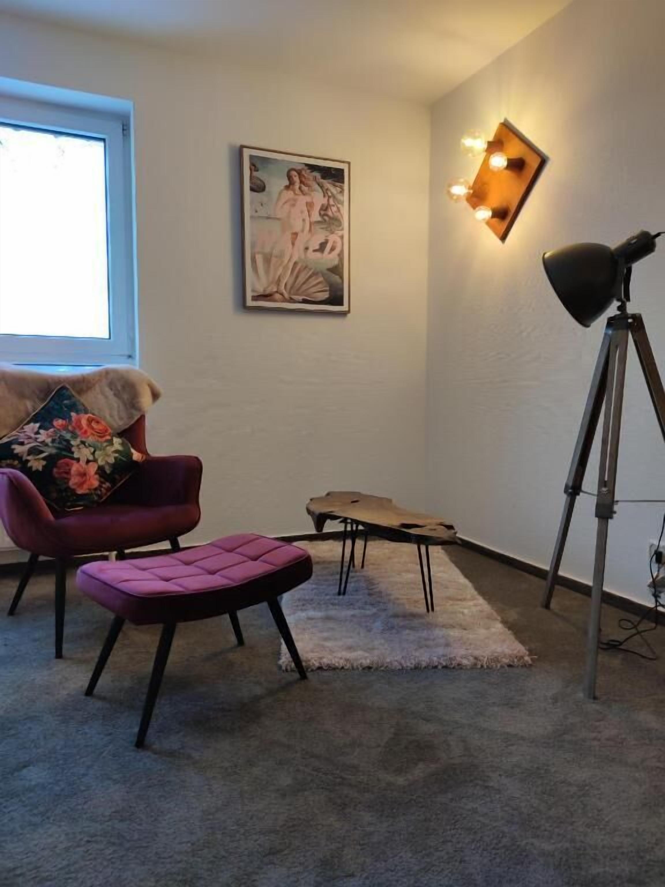 Um quarto com uma cadeira vermelha, um pequeno banquinho, uma mesa de madeira, um abajur em tripé e um quadro na parede.
