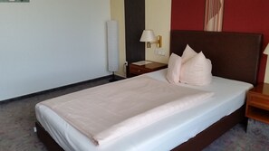 Cama bem arrumada com edredom branco e almofadas, mesa de cabeceira com abajur e luminária fixada na parede.