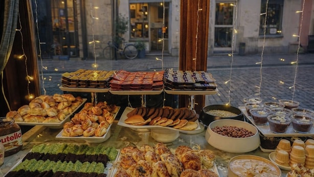 Diversos pastéis e sobremesas expostos numa mesa em frente a um edifício, com uma rua de calçada e edifícios ao fundo.
