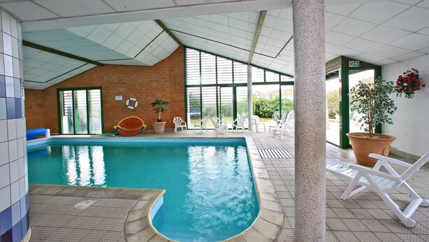 Piscina interna com piso de azulejos, parede de tijolos e teto de vidro. Há cadeiras de descanso brancas e um vaso de planta perto da piscina.