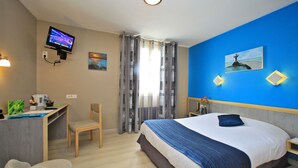 Quarto de hotel com cama, escrivaninha, cadeira, televisão, cortinas e obras de arte nas paredes.