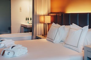 Quarto de hotel com cama, almofadas, mesa de cabeceira, luminária e área de banheiro com pia e artigos de higiene.