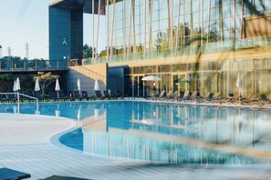 Piscina com espreguiçadeiras e guarda-sóis, cercada por um edifício moderno com grandes janelas.