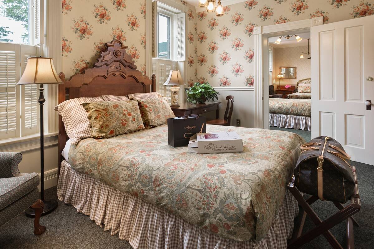 Quarto com papel de parede floral, cabeceira de madeira, cama com roupa de cama florida, mesa de cabeceira, cadeira, abajur e mala.