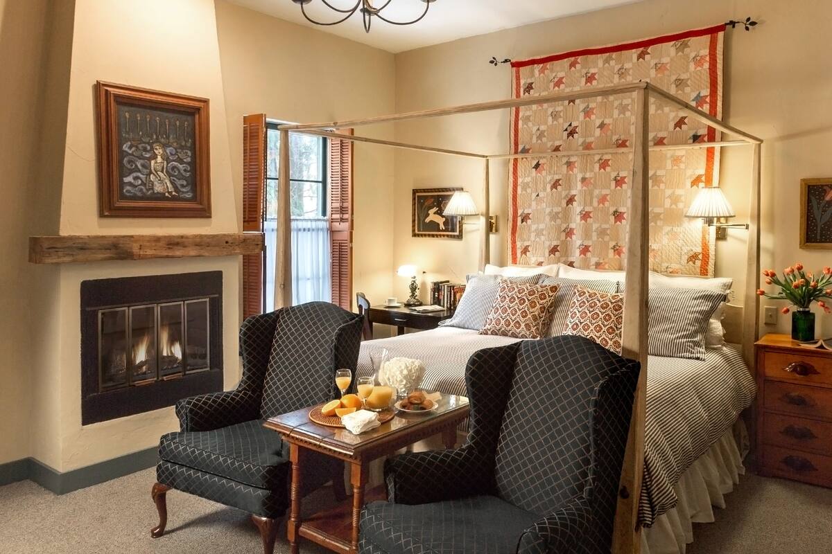 Uma cama com dossel coberta por um edredom floral, uma lareira, duas poltronas e uma mesa com itens de café da manhã.